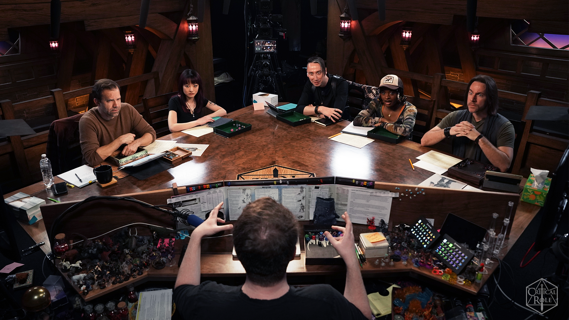 A group of players sit around a wooden table with role playing game character sheets resting in front of them. In the foreground, the Game Master gestures, sort of like a conductor.